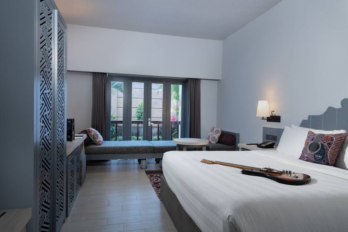 Quarto de hotel com cama, um violão, sofá, uma mesinha e vista do exterior por uma grande janela.