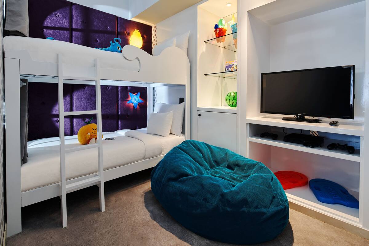 Uma cama de solteiro com estampa de noite estrelada, um puff azul-turquesa, uma TV e prateleiras com brinquedos e jogos.