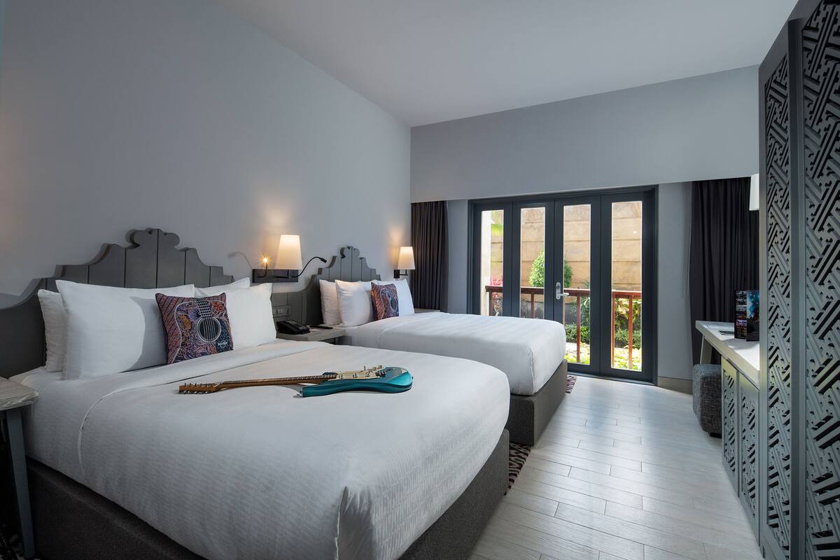 Quarto de hotel com duas camas, um violão, uma escrivaninha e vista para o exterior.