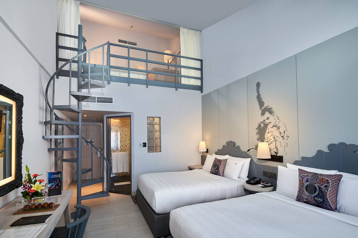 Quarto de hotel com cama de casal, escrivaninha com cadeira, mesinha com um vaso de flores, escada e uma obra de arte na parede.