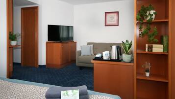 Quarto de hotel com cama, televisão, sofá, mesa de centro, estante e planta.