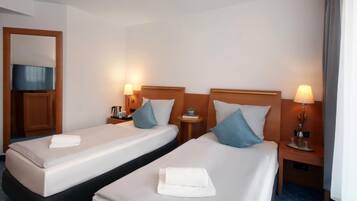 Quarto de hotel com duas camas, cada uma com travesseiro e toalha dobrada. Há uma televisão na parede e uma pequena mesa com abajur.