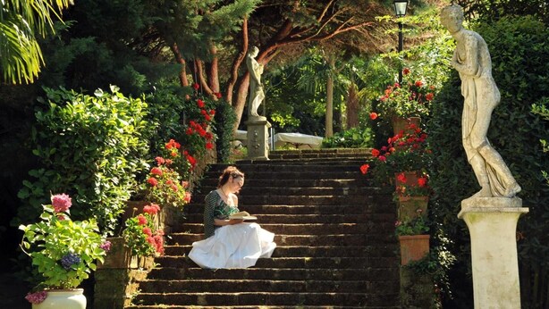 Mulher sentada em degraus de pedra, rodeada por vegetação exuberante e flores vermelhas.