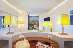 Quarto de hotel com um banco de madeira curvado, vista para a cidade através de uma janela grande, e duas luminárias de cada lado do banco.