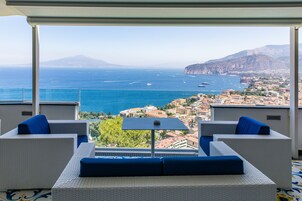 Varanda com móveis de vime e uma mesa, com vista para uma cidade costeira, barcos e montanhas.