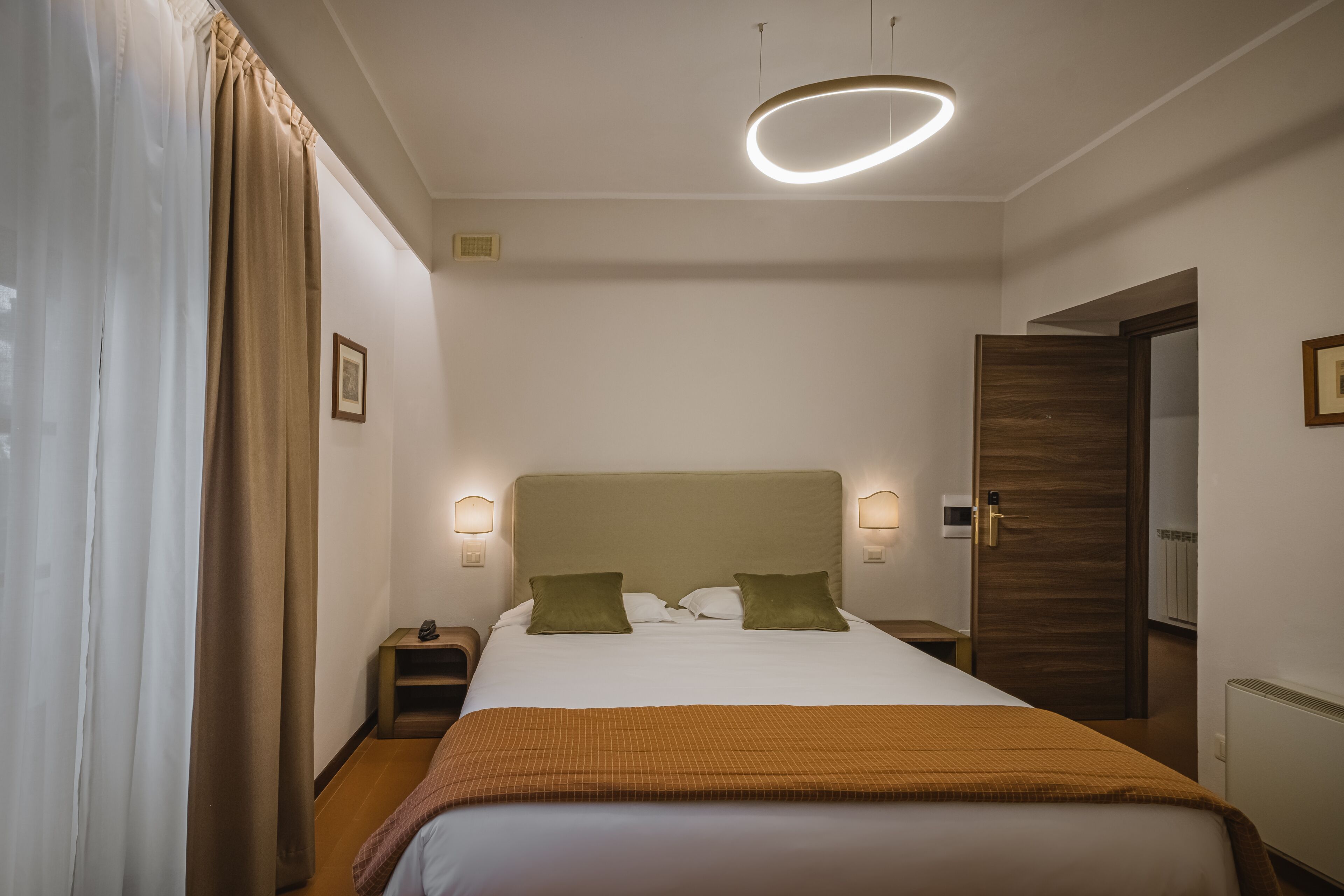 Quarto de hotel com uma cama grande, duas mesas de cabeceira, uma mesinha de apoio, uma porta de madeira e um lustre no teto.