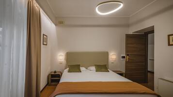 Quarto de hotel com uma cama grande, duas mesas de cabeceira, uma mesinha de apoio, uma porta de madeira e um lustre no teto.