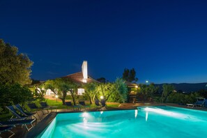 Piscina com espreguiçadeiras, um edifício bem iluminado e um céu noturno claro.