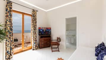 Quarto com televisão, uma cadeira de madeira, cama e vista para o mar através de uma porta de vidro.
