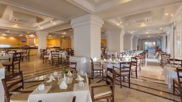Uma ampla sala de jantar com várias mesas e cadeiras, uma cozinha ao fundo e vista para o exterior através de grandes janelas.