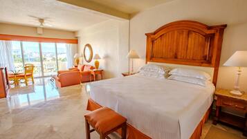 Quarto de hotel com uma cama grande, cabeceira de madeira, área de estar com uma cadeira e mesa, e varanda com vista.