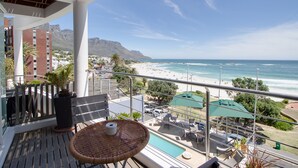 Varanda com vista para uma praia e oceano, com piscina, espreguiçadeiras e guarda-sóis.