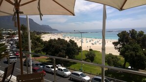 Vista para a praia a partir de uma varanda, com o oceano, guarda-sóis e carros estacionados.