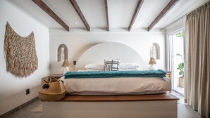 Quarto com uma cama grande, vigas de madeira, arco na parede e um enfeite de macramé pendurado.
