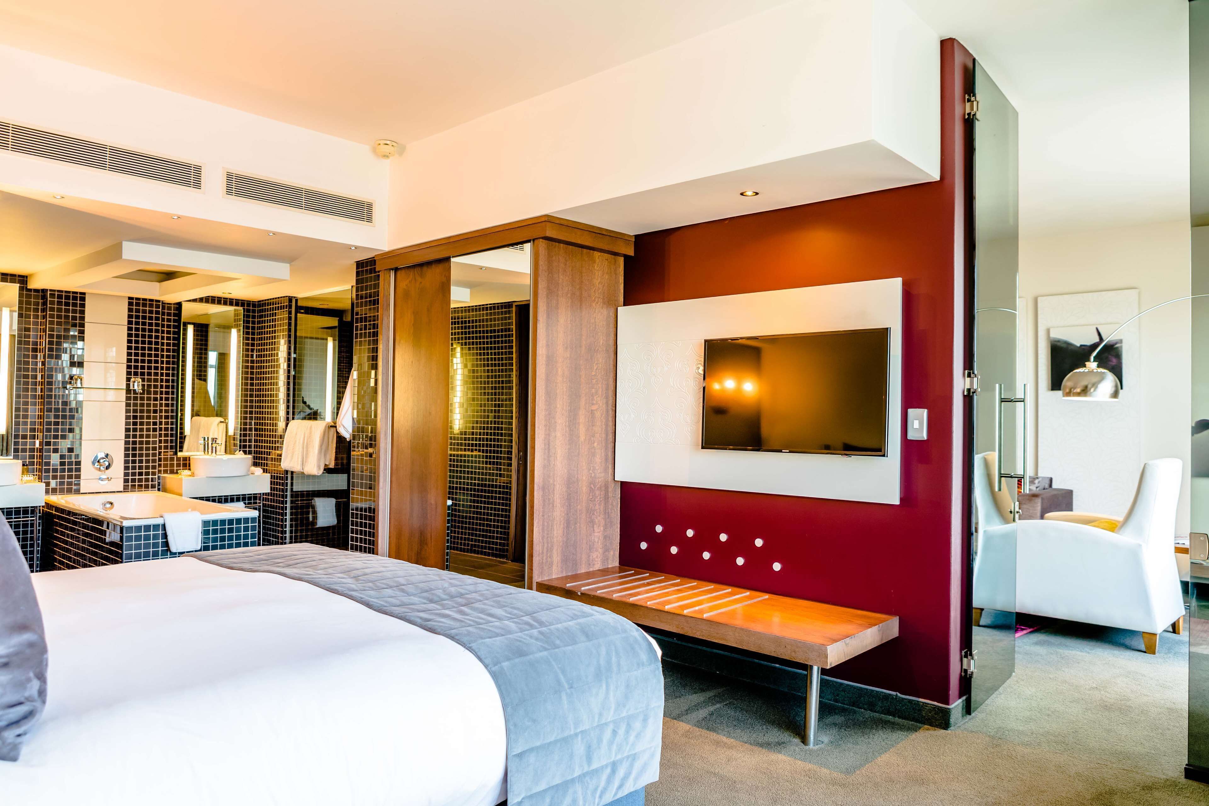 Quarto de hotel moderno com uma cama grande, uma penteadeira com espelho, área de estar e uma televisão fixada na parede.