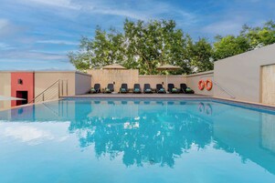 Piscina com espreguiçadeiras e guarda-sóis, cercada por um muro baixo e árvores.