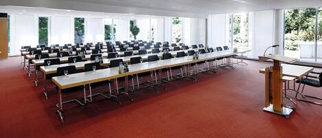 Sala de conferências com mesas e cadeiras dispostas em fileiras, janelas amplas e um palanque.