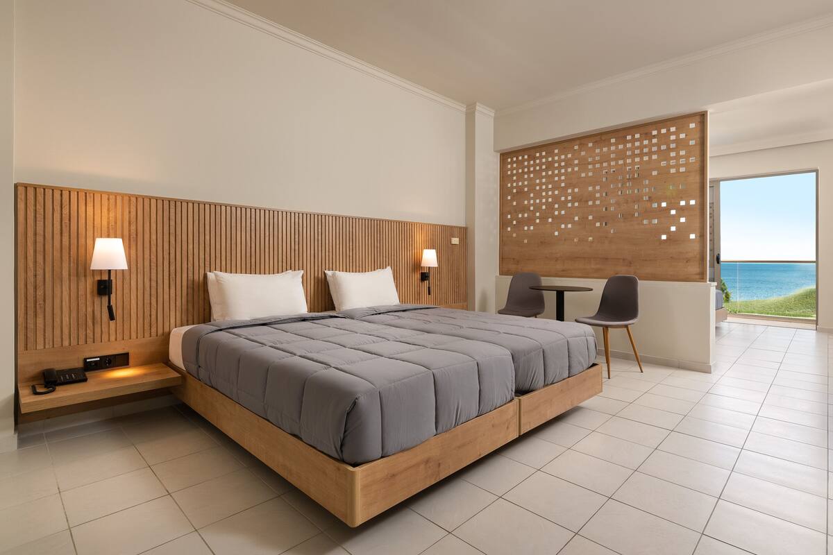 Quarto de hotel moderno com uma cama grande, luminárias de cabeceira, uma mesinha e vista para o mar.