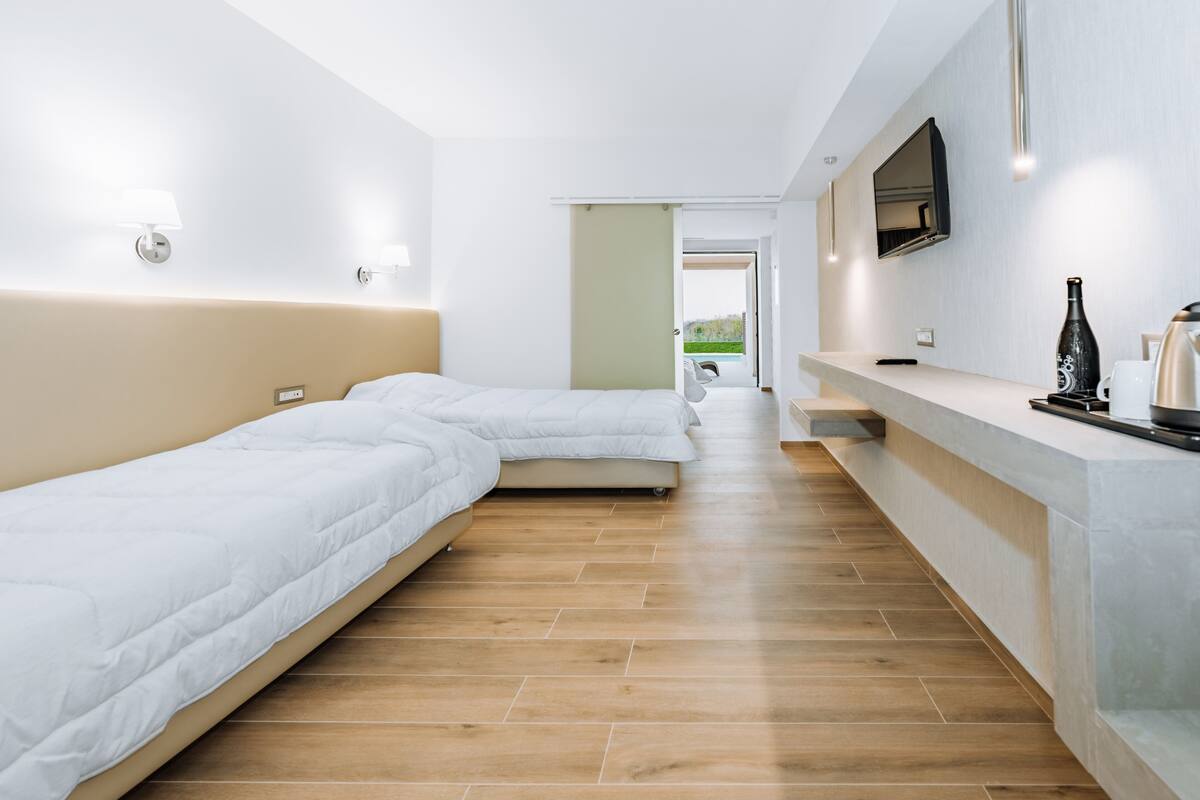 Quarto de hotel moderno com uma cama grande, um sofá pequeno, uma TV na parede e piso de madeira.