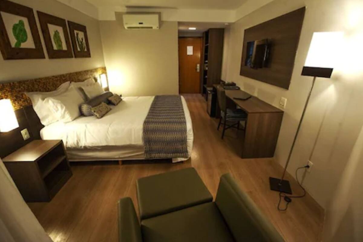 Quarto de hotel com cama, escrivaninha, cadeira e televisão na parede.