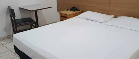 Uma cama de solteiro com cabeceira de madeira, uma mesinha de madeira e uma cadeira em um quarto com piso de azulejos.