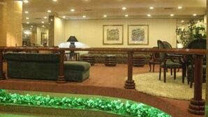 Um hall de hotel com um sofá verde, uma área de refeições com cadeiras, um abajur e obras de arte emolduradas na parede.