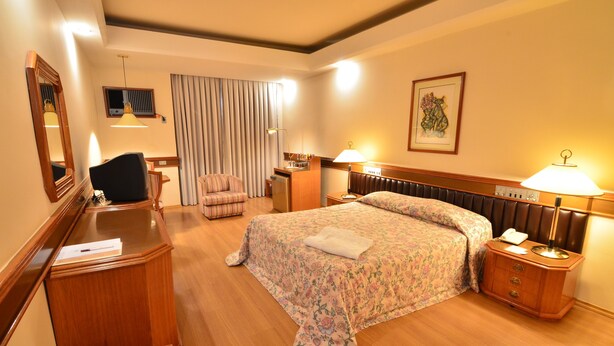 Quarto de hotel com cama, escrivaninha, cadeira, mesa de cabeceira e espelho.