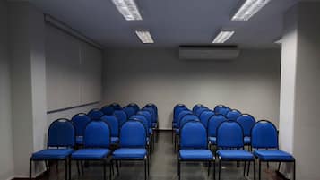 Uma sala de conferências com fileiras de cadeiras azuis, uma tela de projetor e luminárias no teto.