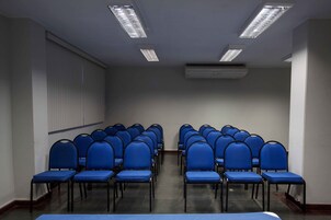Uma sala de conferências com fileiras de cadeiras azuis, uma tela de projetor e luminárias no teto.