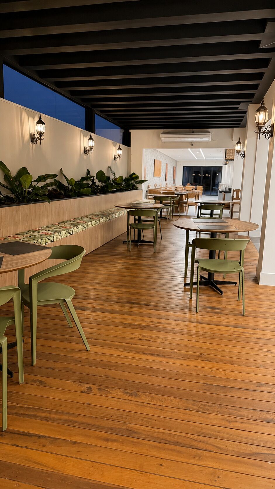 Restaurante com piso de madeira, cadeiras verdes e mesas. Há plantas ao longo da parede e luminárias fixadas na parede.