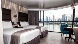 Quarto de hotel com duas camas, uma escrivaninha e uma janela grande com vista para a cidade.