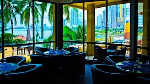 Restaurante com grandes janelas que oferecem vista para o horizonte urbano e palmeiras.