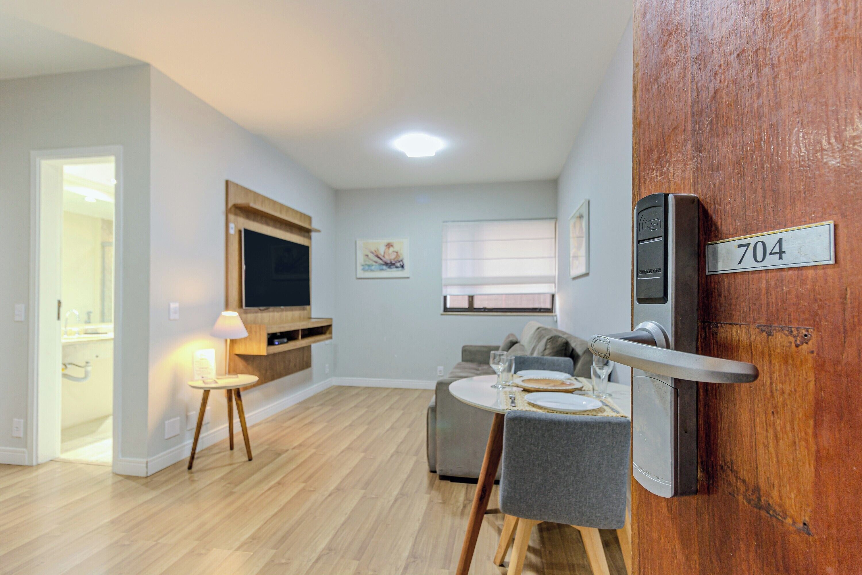 Uma sala de estar moderna com televisão de tela plana, uma área de jantar com mesa posta para dois e uma porta de madeira com placa de identificação.