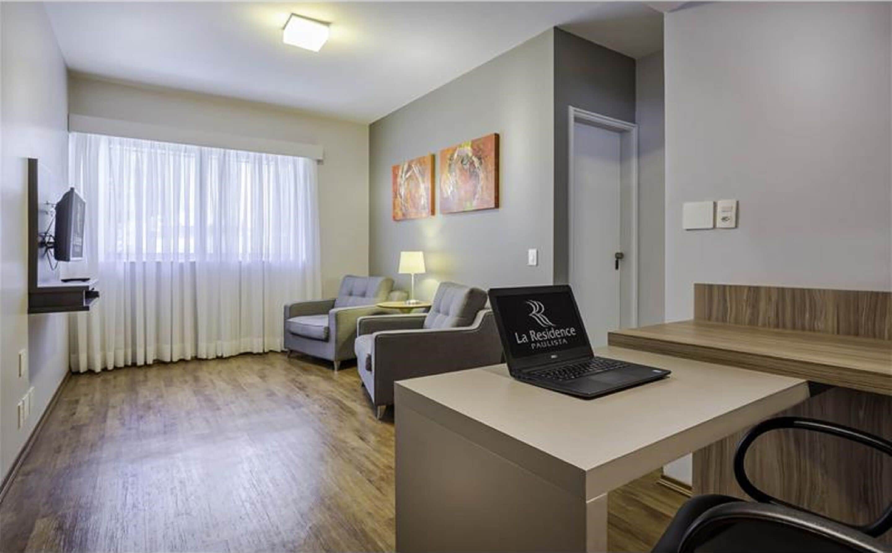 Quarto de hotel com escrivaninha, duas cadeiras, televisão, luminária e obras de arte na parede.