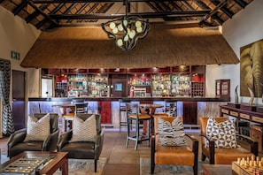 Uma área de bar com assentos de couro, teto de palha e uma variedade de garrafas em prateleiras.