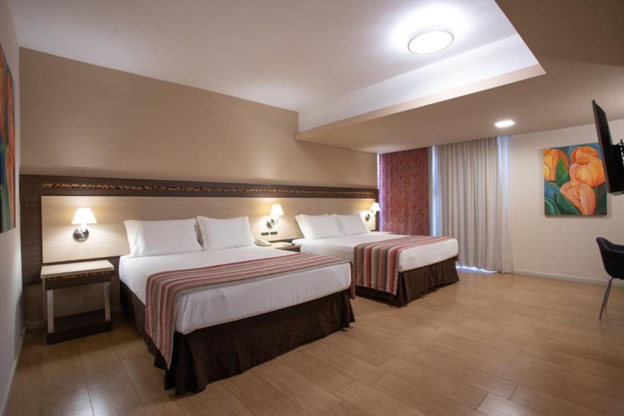 Quarto de hotel com duas camas, uma escrivaninha, uma televisão e um quadro na parede.