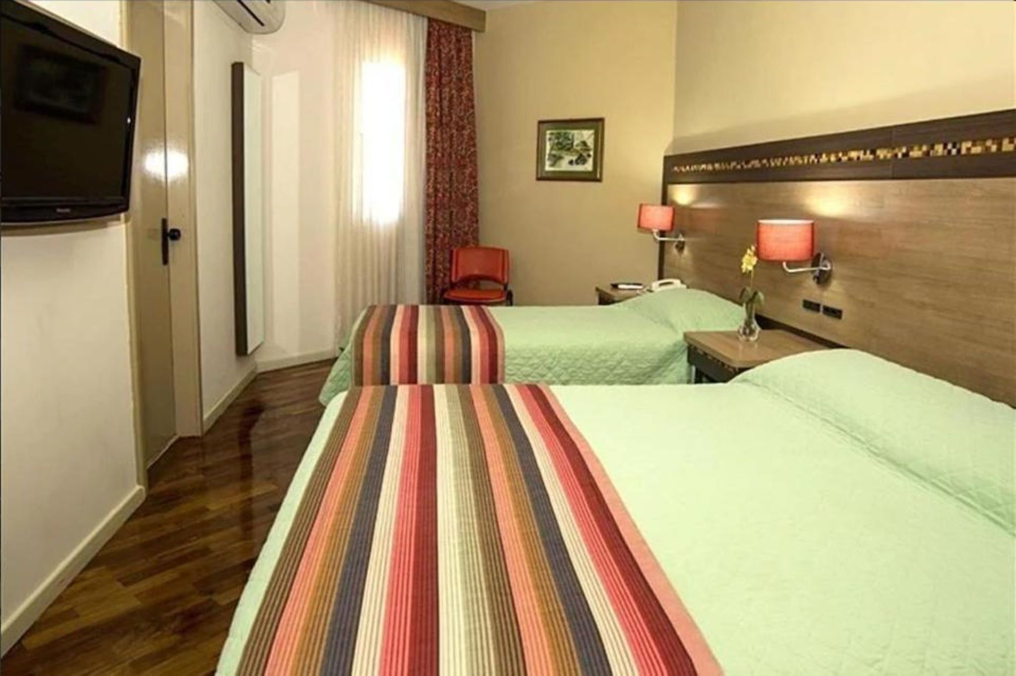 Quarto de hotel com duas camas, televisão, uma cadeira, escrivaninha e luminária na parede.