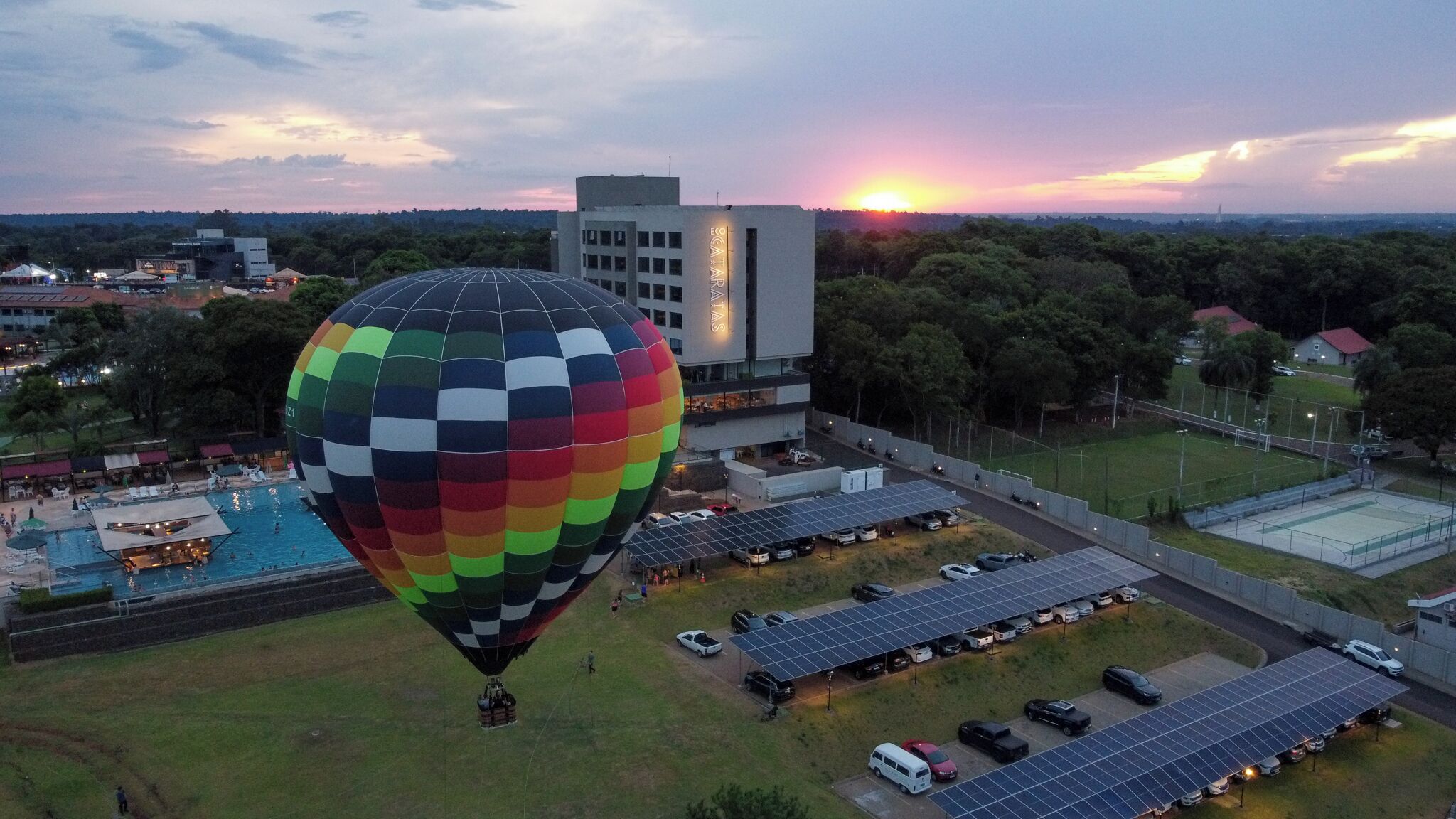 Um balão de ar quente sobre uma área de resort com piscina, painéis solares e um edifício com a placa 'ECATAS'.