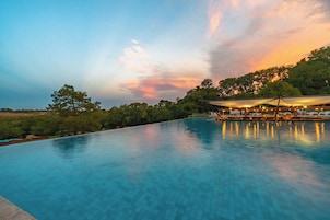 Piscina com vista para o pôr do sol, rodeada por vegetação e área de estar coberta com iluminação.