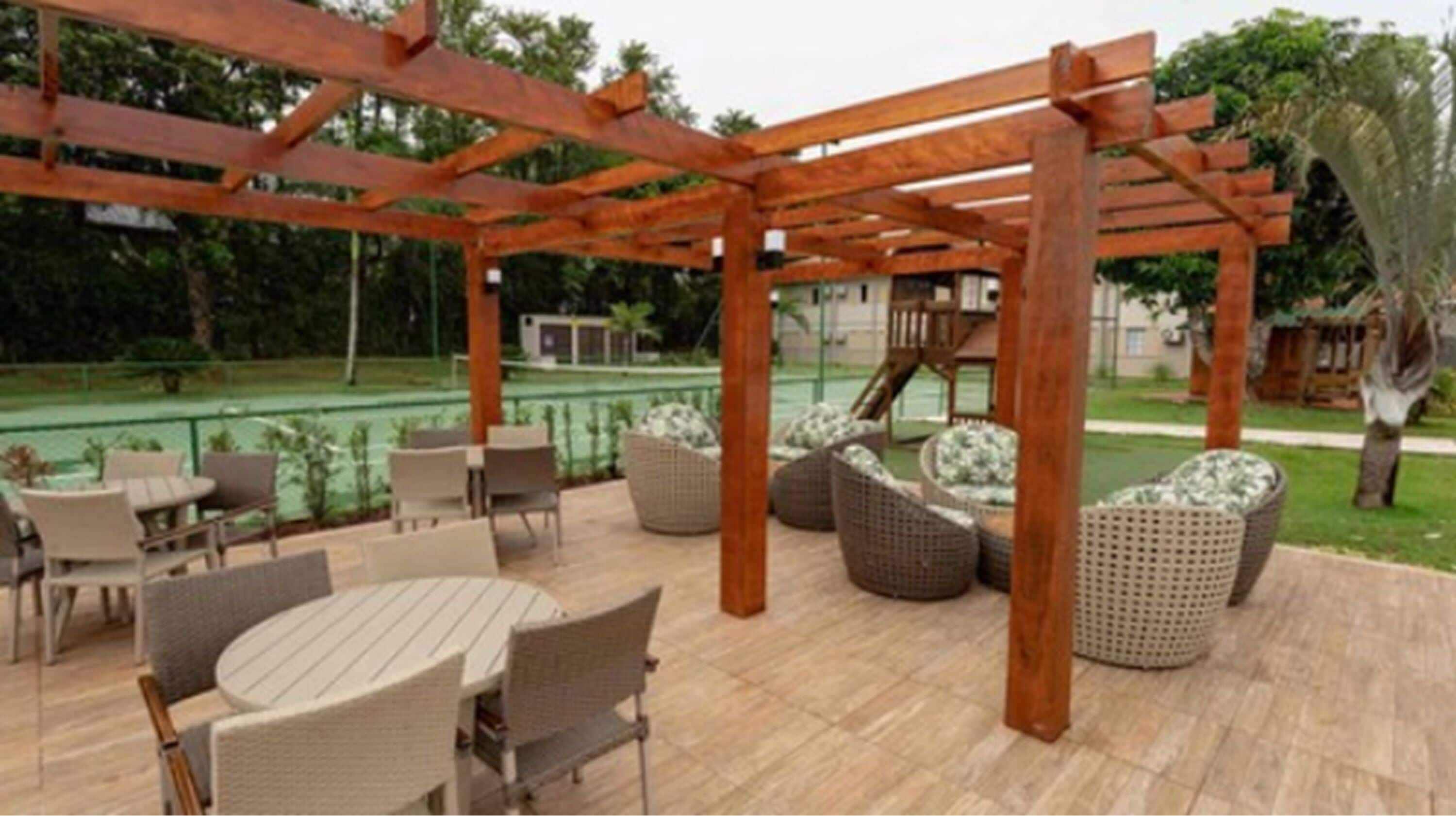 Pátio externo com pérgola de madeira, mobiliário de vime e vista para uma piscina.