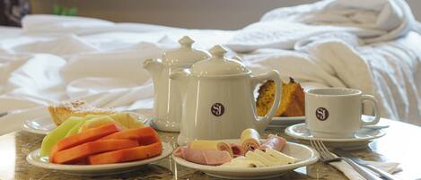 Café da manhã servido na cama, com pratos de fruta, queijo e presunto, um bule, xícara de café e talheres.