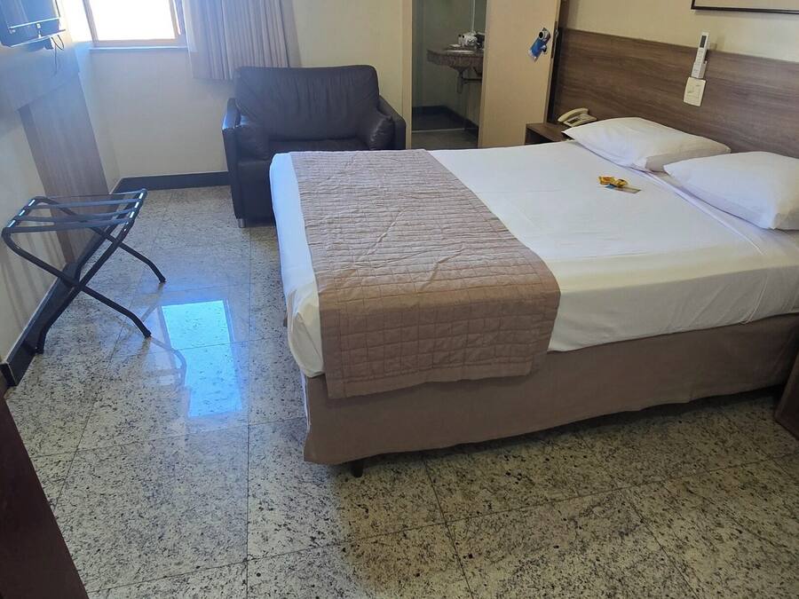 Quarto de hotel com cama, uma cadeira, uma escrivaninha e um banheiro visível por uma porta aberta.
