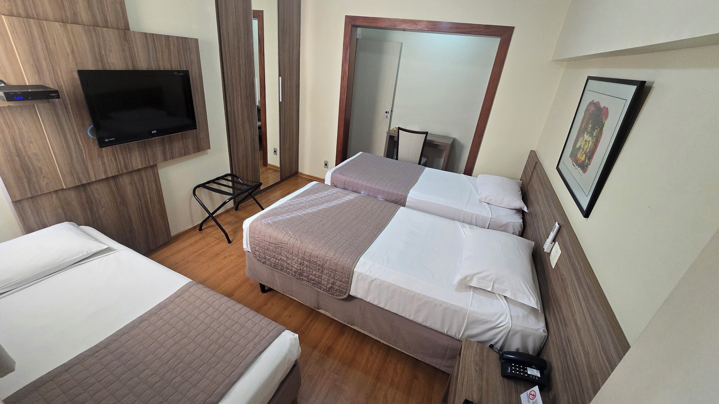 Quarto de hotel com duas camas, uma televisão na parede, uma mesinha e um quadro pendurado na parede.