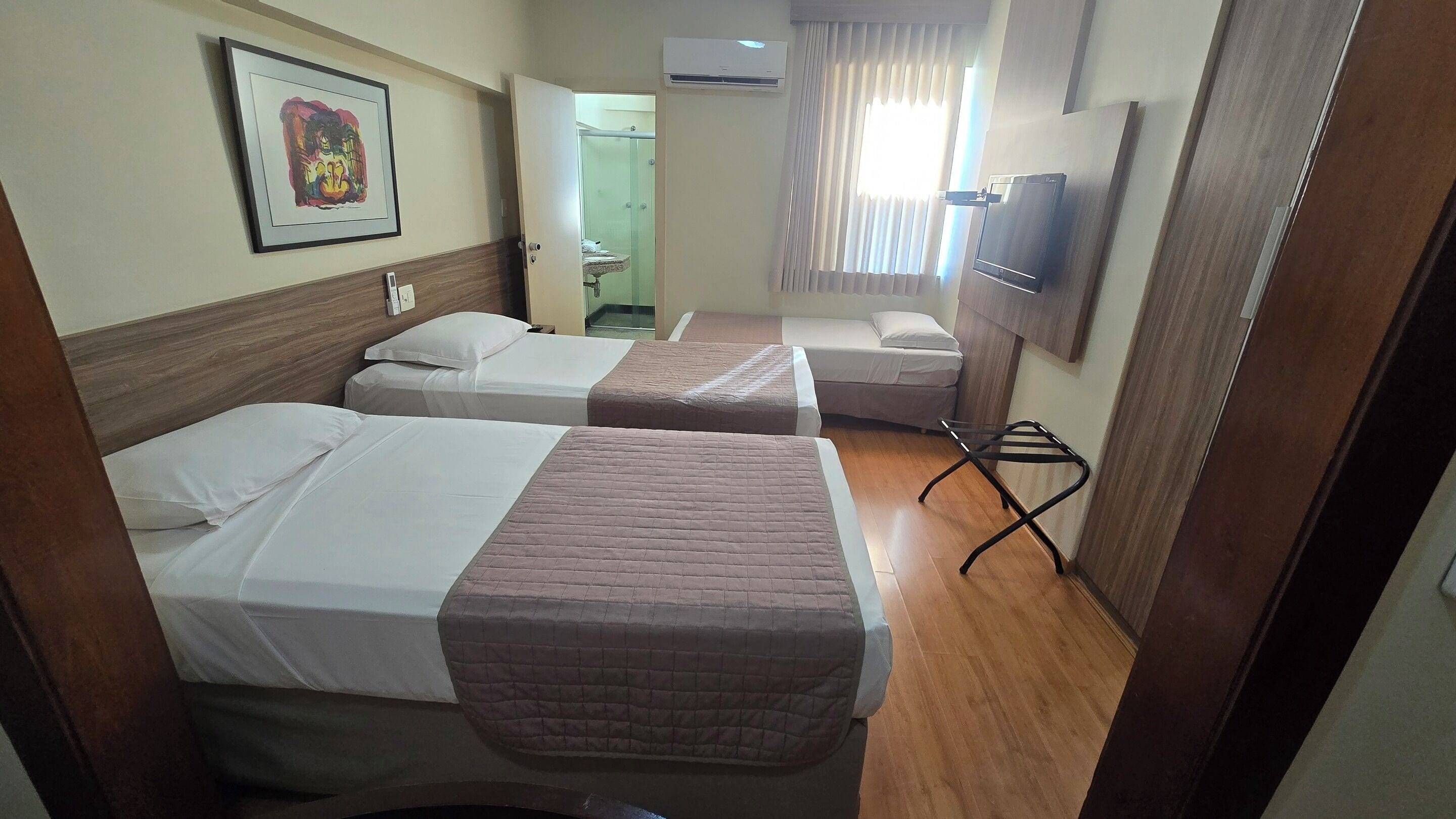 Quarto de hotel com duas camas, uma mesa pequena, uma televisão e um banheiro visível por uma porta aberta.