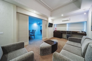 Uma sala de estar moderna com sofás estofados cinza, uma mesa de centro escura e uma área de cozinha com televisão e aparelho de ar condicionado.