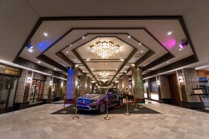 Um saguão de hotel luxuoso com um lustre central, um carro Mercedes-Benz em exposição e uma barreira de corda vermelha.