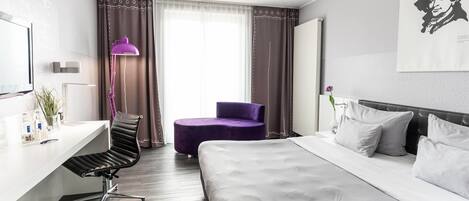 Quarto de hotel com cama, escrivaninha, cadeira, um banquinho roxo, luminária e um quadro na parede.