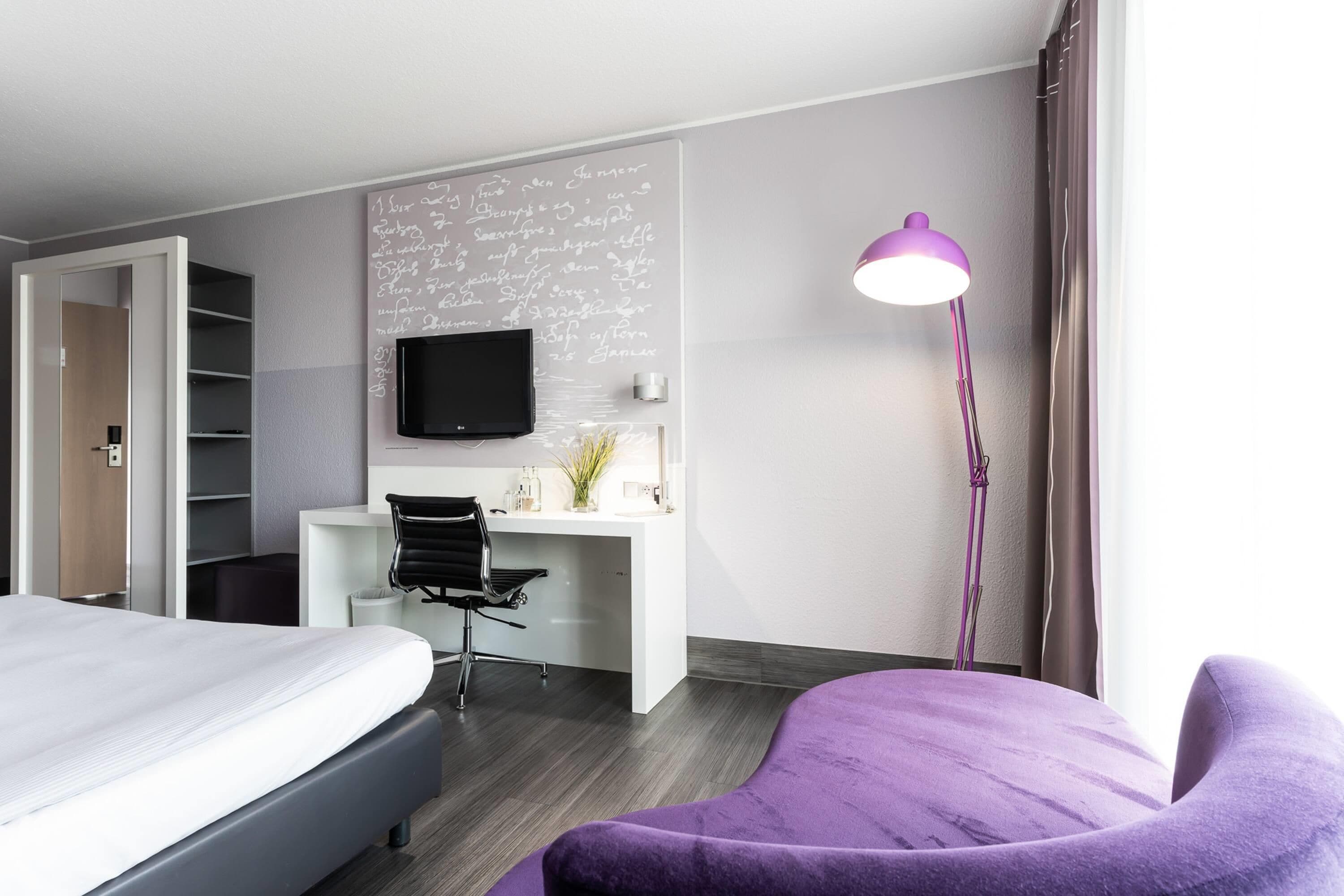 Quarto de hotel com uma cama, uma escrivaninha com cadeira, uma televisão, uma poltrona roxa e um abajur.