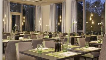Uma sala de jantar com mesas preparadas para vários convidados, amplas janelas com cortinas e luminárias pendentes.