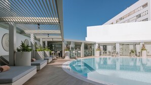 Uma área de piscina moderna, com assentos, uma piscina azul límpida e um edifício com grandes janelas.